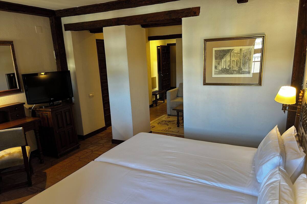 Quarto de hotel com uma cama grande, televisão sobre um móvel de madeira, quadro na parede e uma luminária.