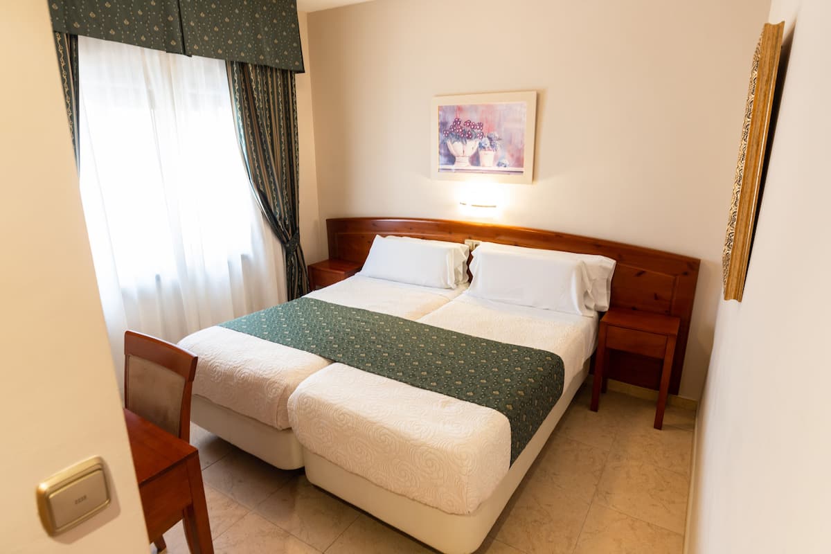 Uma cama de casal com cabeceira de madeira, duas mesas de cabeceira, um edredom verde estampado e um quadro na parede.