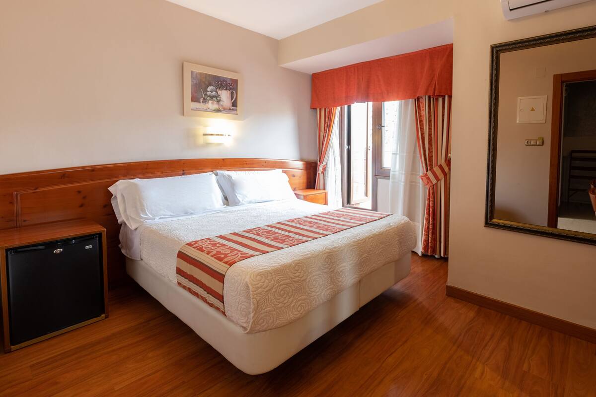 Quarto de hotel com cama de cabeceira em madeira, pequena geladeira, mesa de cabeceira e janela com cortinas.