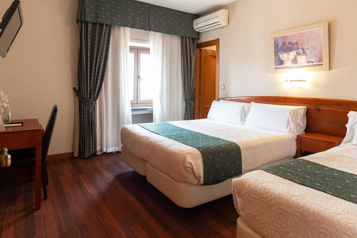Quarto de hotel com duas camas, piso de madeira, uma mesa de trabalho com cadeira e um quadro na parede.