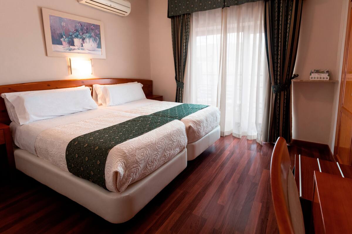 Quarto de hotel com uma cama grande, cabeceira de madeira, mesa de cabeceira, escrivaninha e uma cadeira.