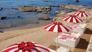 Uma praia com águas azul-claras, costa rochosa e guarda-sóis listrados em vermelho e branco.