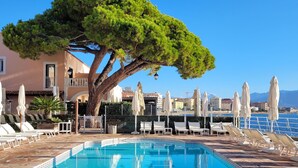 Uma área de piscina com uma grande árvore, cadeiras de descanso e guarda-sóis.