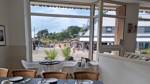 Uma sala de jantar com mesa posta para quatro, amplas janelas com vista para uma praça pública com pessoas e áreas verdes.