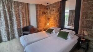 Quarto de hotel com duas camas, uma parede de pedra, uma cadeira, uma mesa e um abajur.