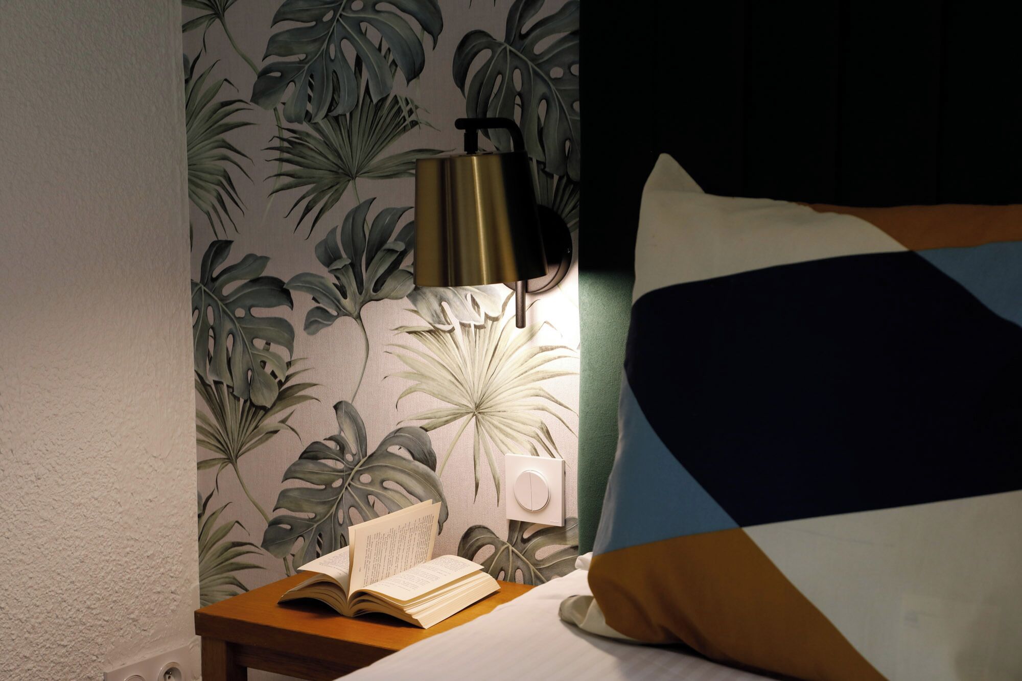 Cama bem arrumada com travesseiro, mesa de cabeceira com um livro e abajur, e parede com padrão de folha tropical.