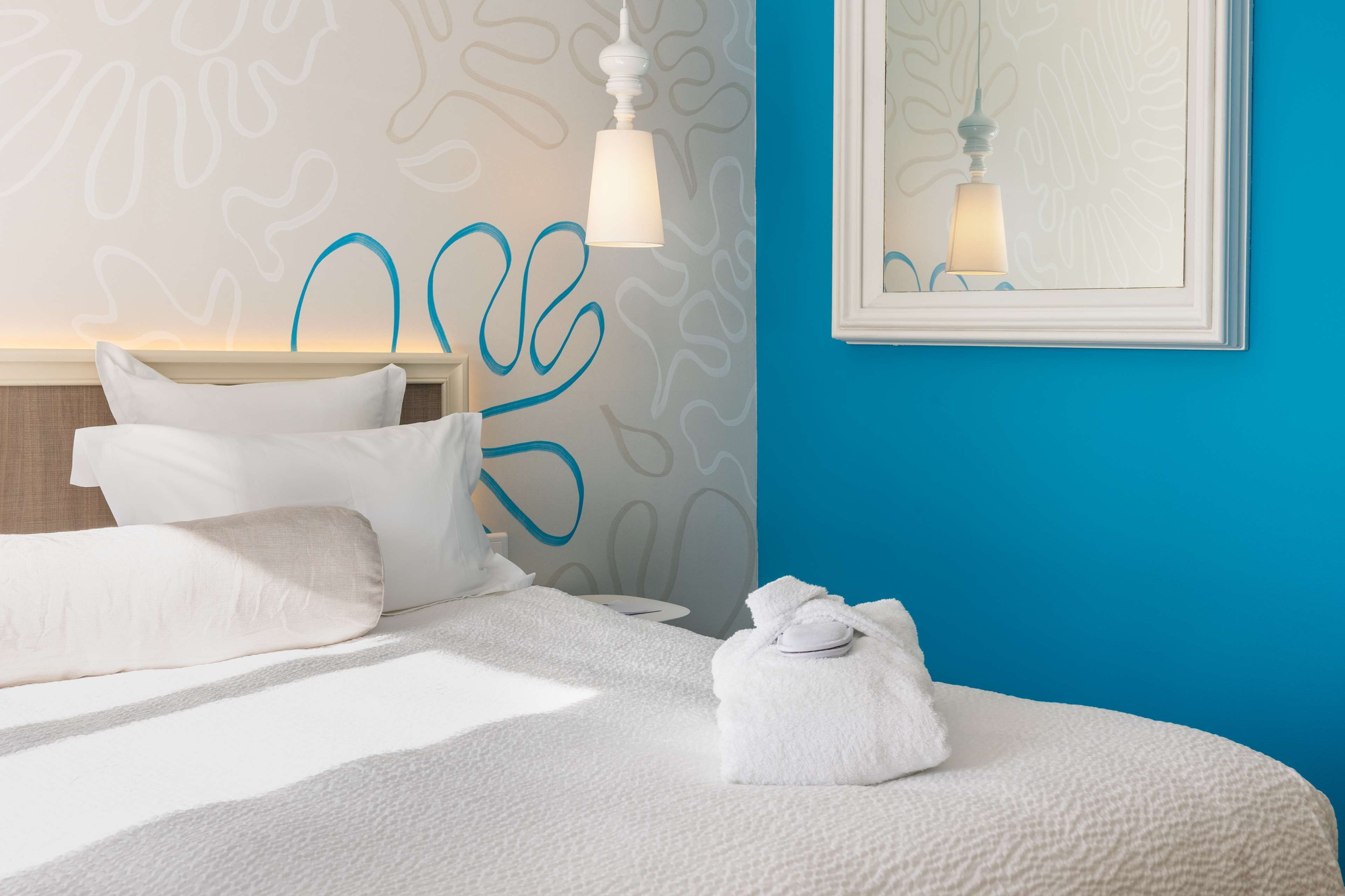 Uma cama bem arrumada com lençóis e almofadas brancas, uma toalha ao lado e uma parede com padrões abstratos em azul e cinza.