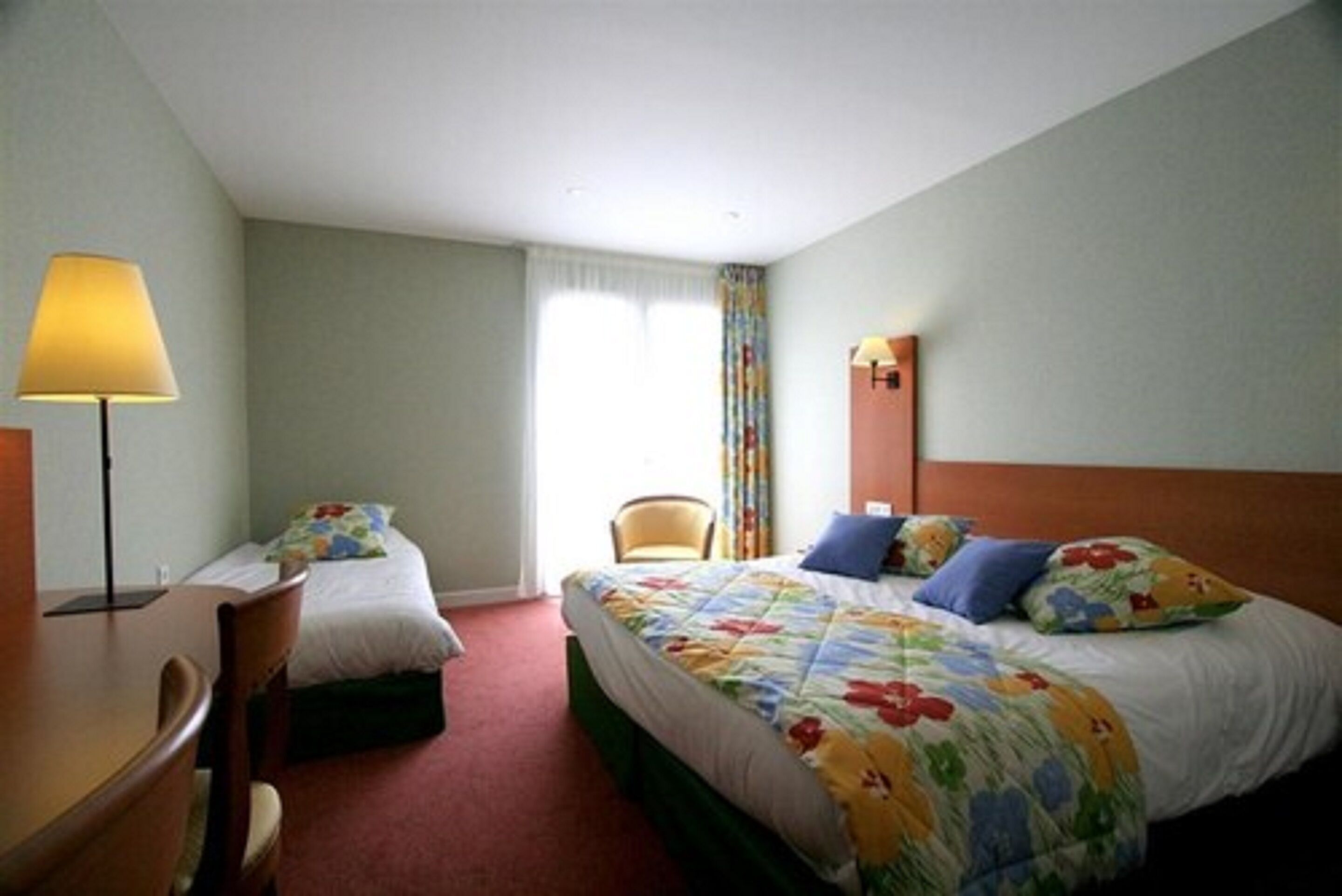 Quarto de hotel com uma cama, uma escrivaninha, uma cadeira, um abajur e uma janela com cortinas.