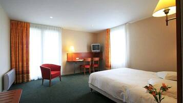 Quarto de hotel com cama, escrivaninha, cadeira, televisão e um vaso com flores.