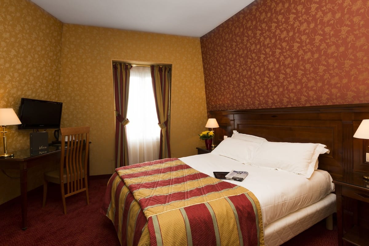 Quarto de hotel com cama, escrivaninha com televisão, cadeira e janela com cortinas.