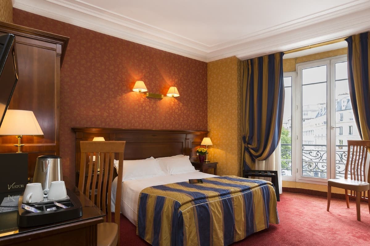 Quarto de hotel com cama, duas luminárias de cabeceira, escrivaninha com cafeteira, cadeira e vista da cidade através de uma janela grande.