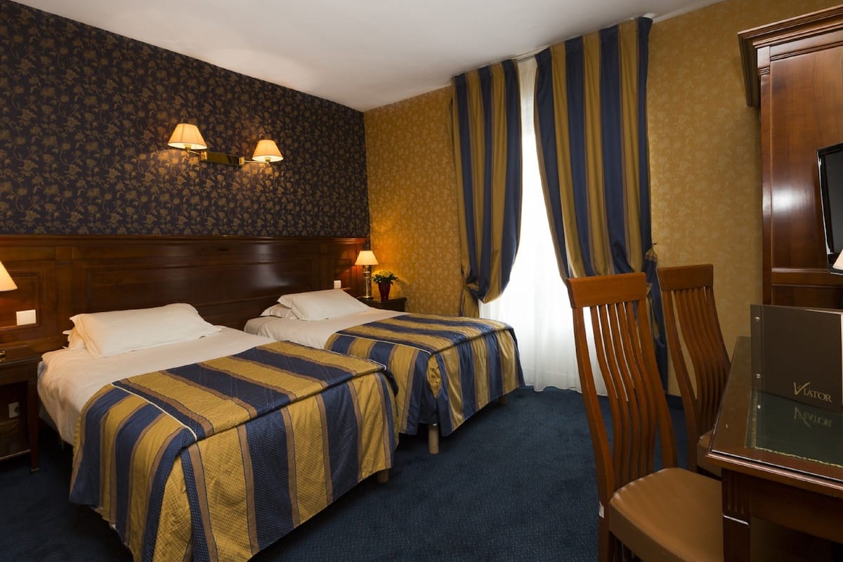 Quarto de hotel com duas camas, uma cabeceira de madeira, uma escrivaninha com cadeira, televisão e uma janela com cortinas.