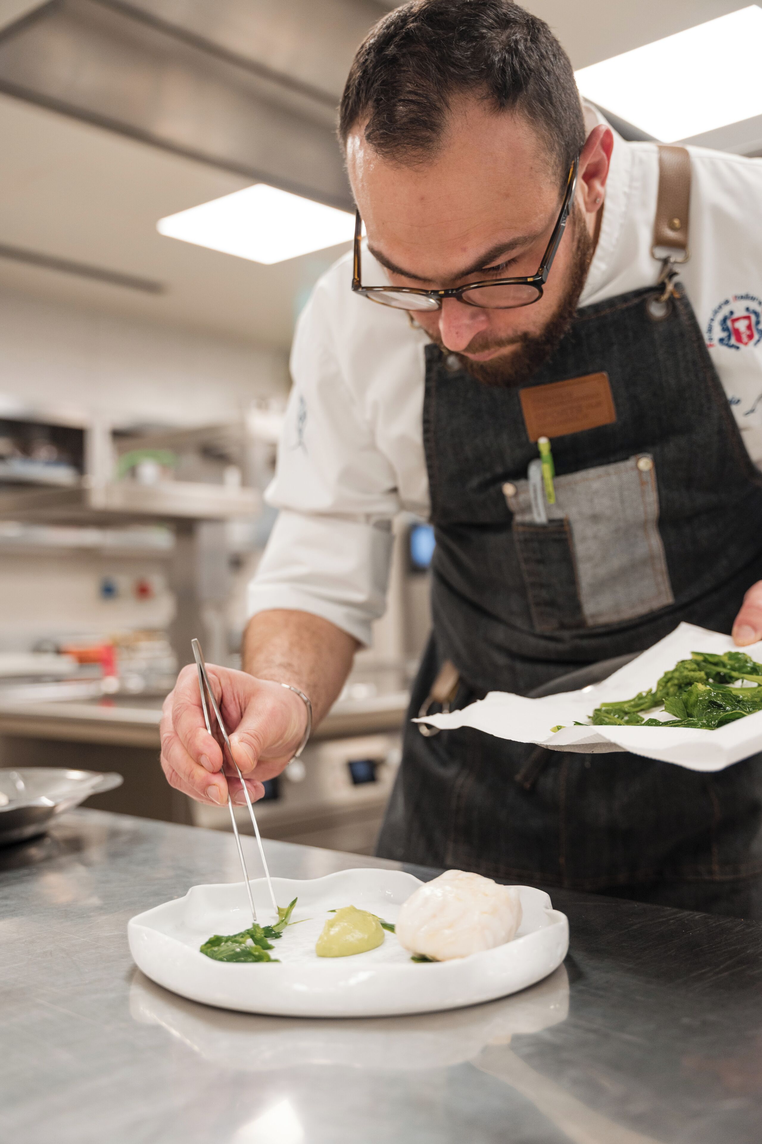 Um chef prepara comida em uma cozinha com bancadas de aço inoxidável e um prato branco com alimentos.