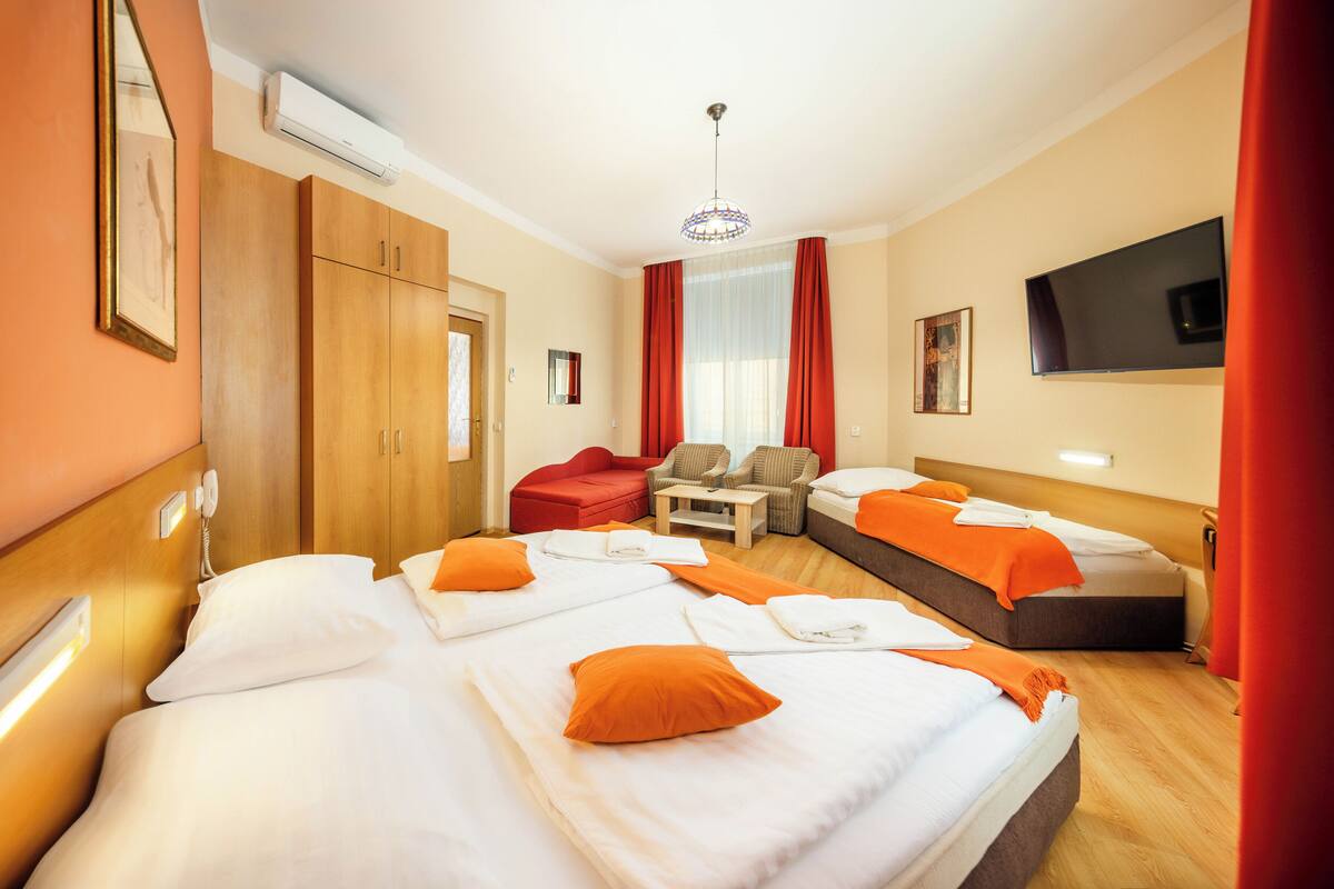 Quarto de hotel com duas camas, um sofá, uma televisão e uma cortina vermelha.