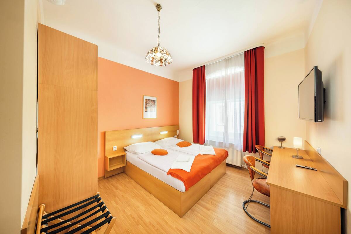 Quarto de hotel com cama de madeira, escrivaninha, cadeira, televisão e janela com cortinas.