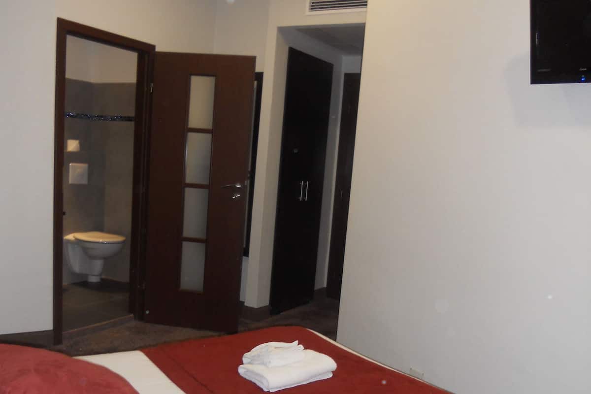 Quarto de hotel com uma cama, um edredom vermelho e branco, um travesseiro branco e uma toalha dobrada sobre a cama. Há uma porta que leva a um banheiro com vaso sanitário e espelho.