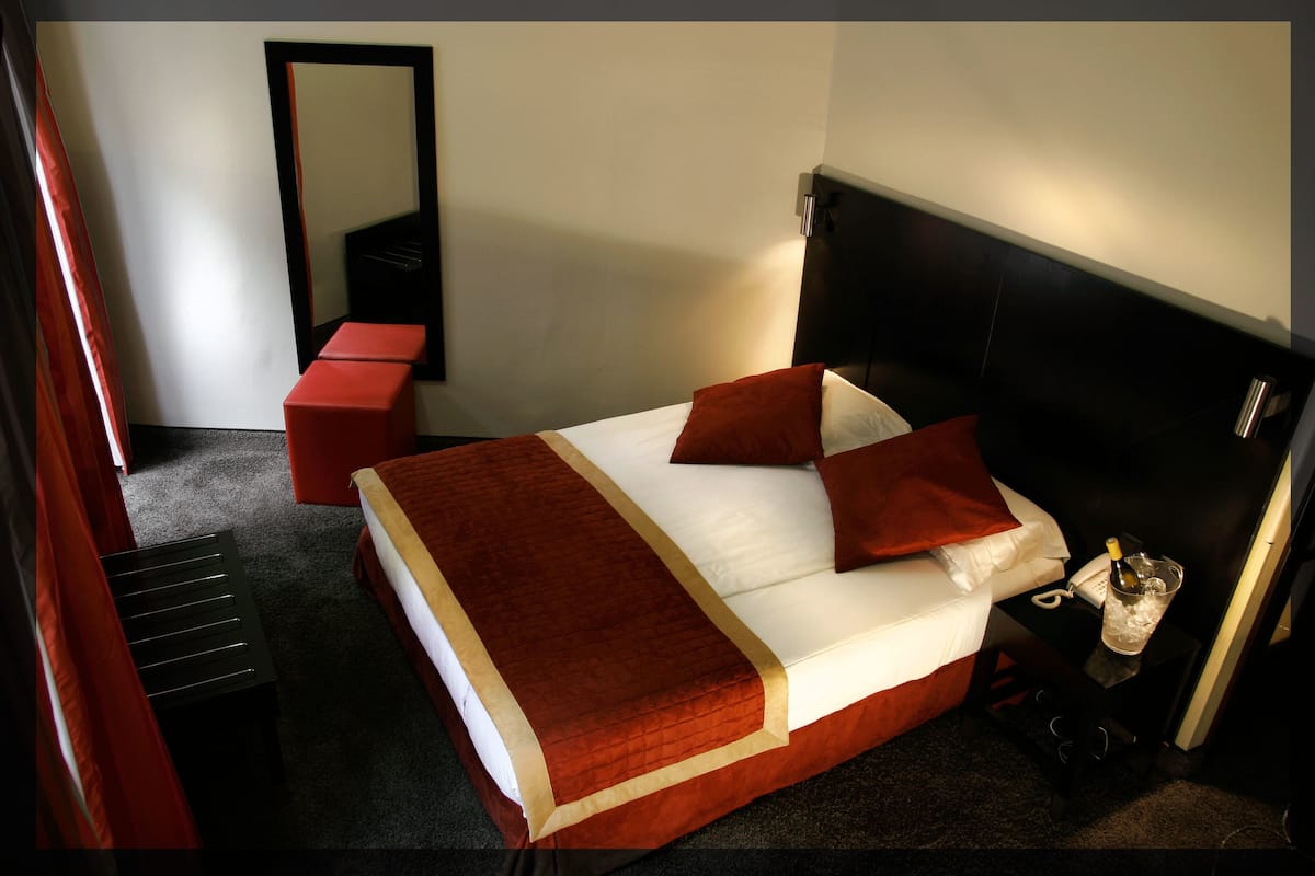 Cama bem arrumada com colcha vermelha e branca, uma pequena mesa de cabeceira com uma garrafa e copos, e um banco vermelho em um quarto de hotel.