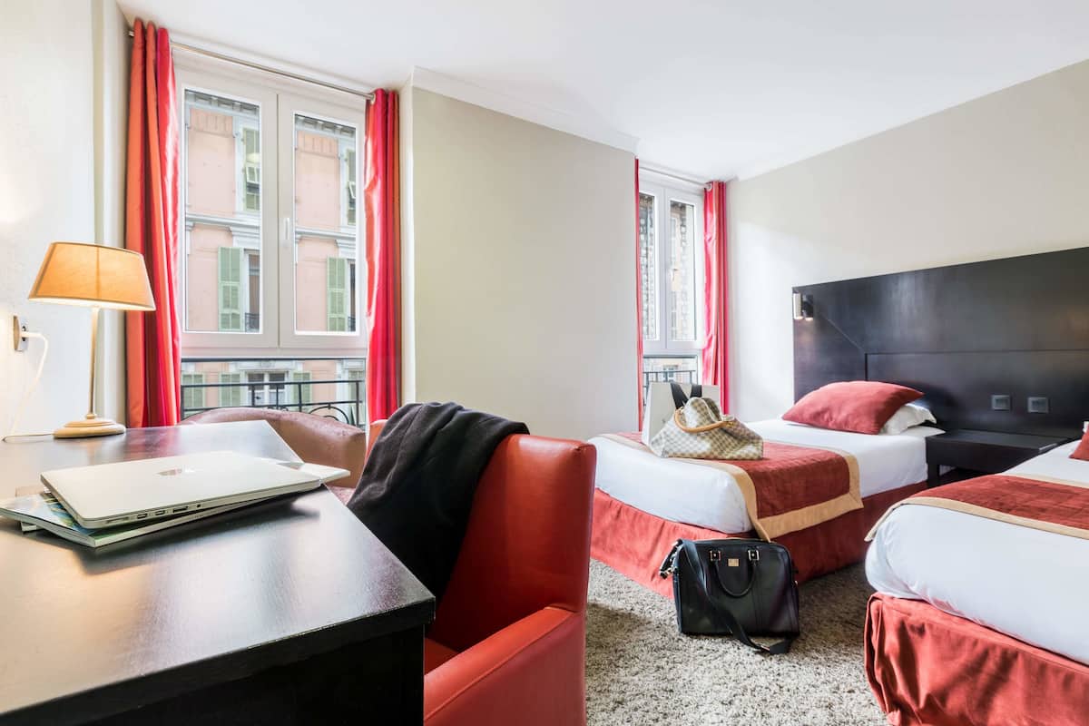 Quarto de hotel com duas camas, uma mesa com um laptop, uma cadeira e uma janela com cortinas vermelhas.