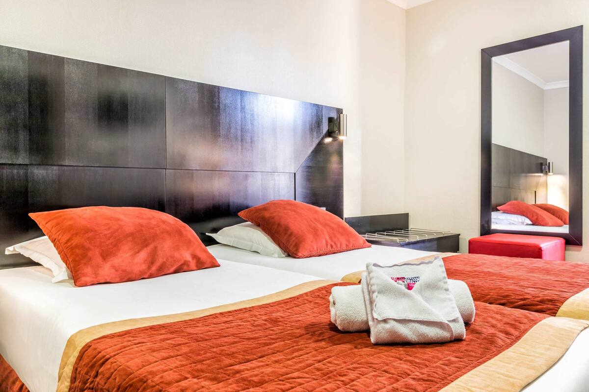 Quarto de hotel com duas camas, roupa de cama vermelha e branca, um espelho grande e um banquinho vermelho.