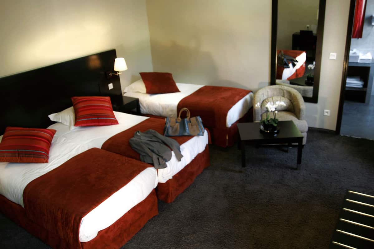 Quarto de hotel com duas camas, cada uma com roupa de cama vermelha e branca, uma pequena mesa com abajur e uma cadeira.