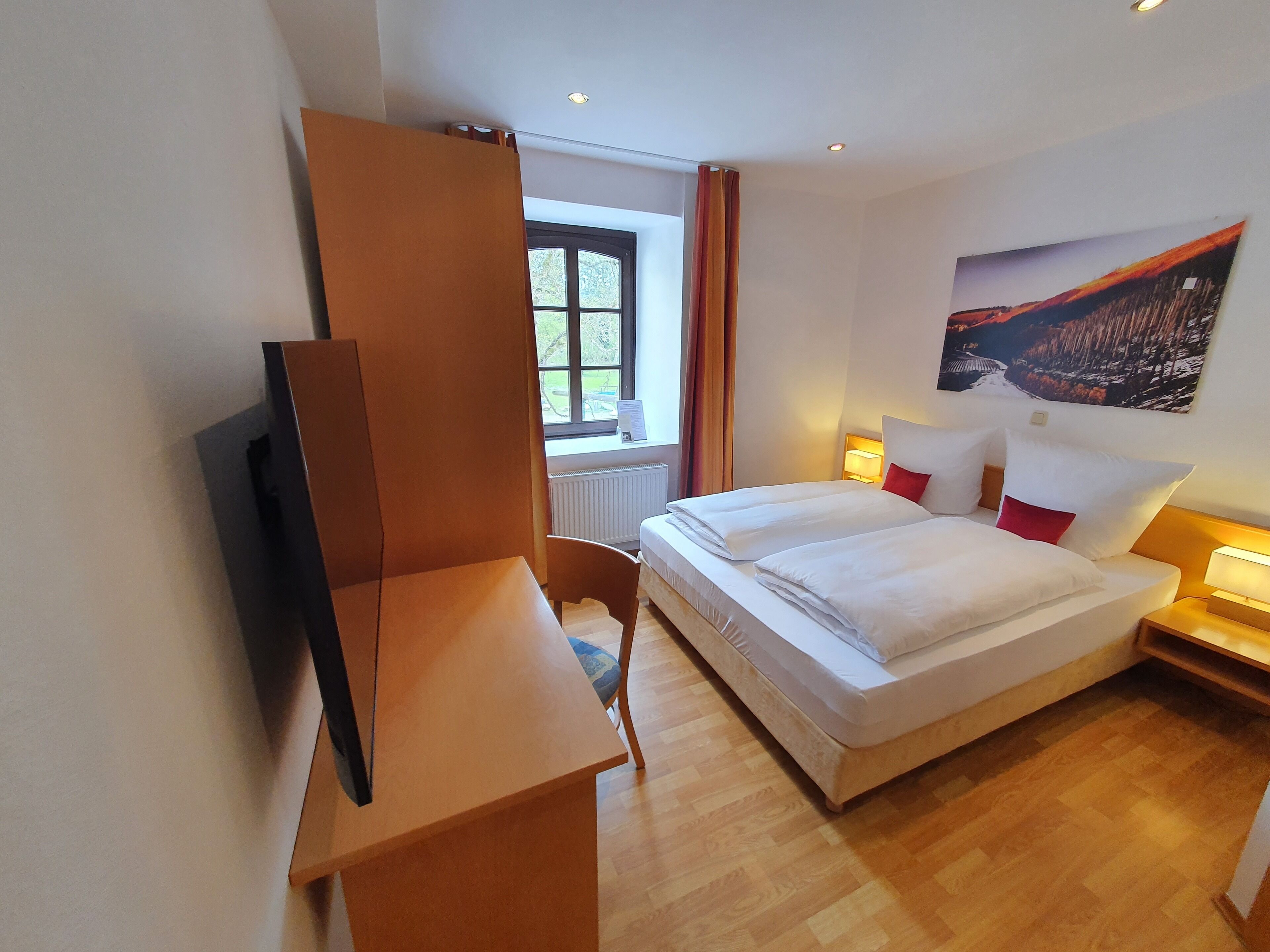 Quarto de hotel com cama, escrivaninha, cadeira, janela com cortinas, um quadro na parede e abajur na mesa de cabeceira.