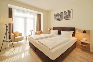 Quarto de hotel com duas camas, uma escrivaninha, uma cadeira e uma janela grande com cortinas.
