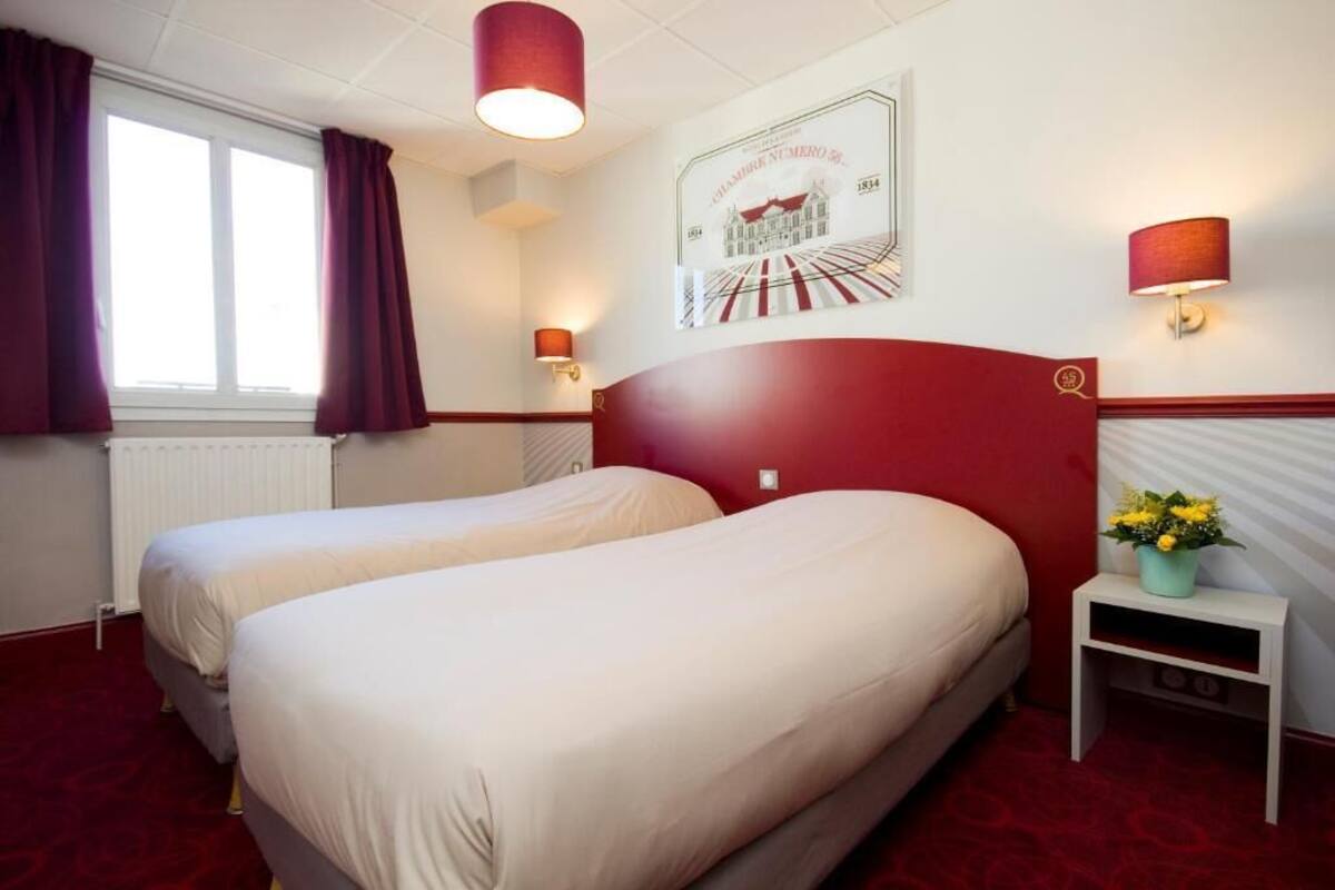 Quarto de hotel com duas camas, uma parede de destaque vermelha, uma janela com cortinas, uma luminária fixada na parede e um quadro emoldurado.