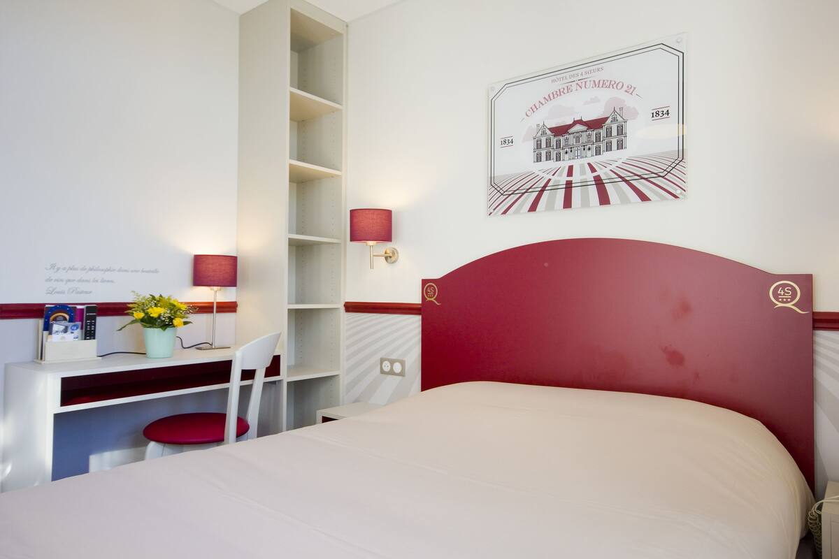 Quarto de hotel com cabeceira vermelha, uma escrivaninha com abajur, uma cadeira e um quadro na parede de um edifício.