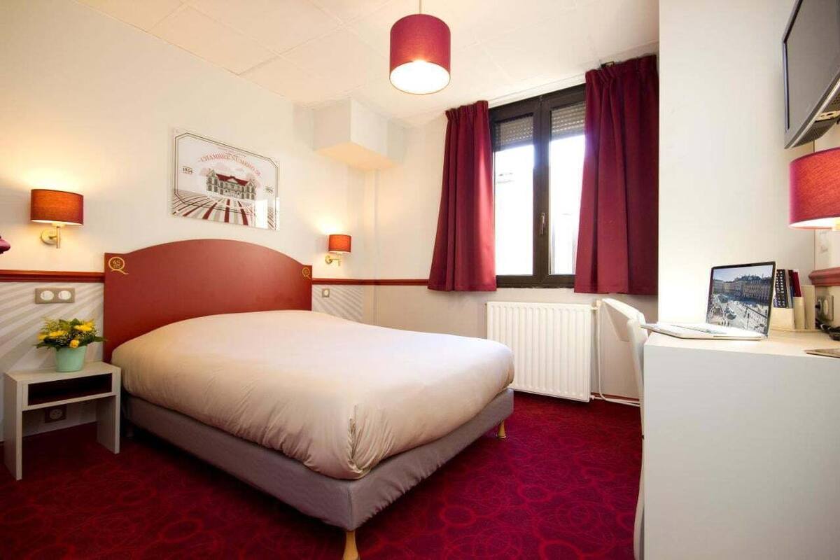 Quarto de hotel com cama, escrivaninha, luminária, radiador e um quadro pendurado na parede.