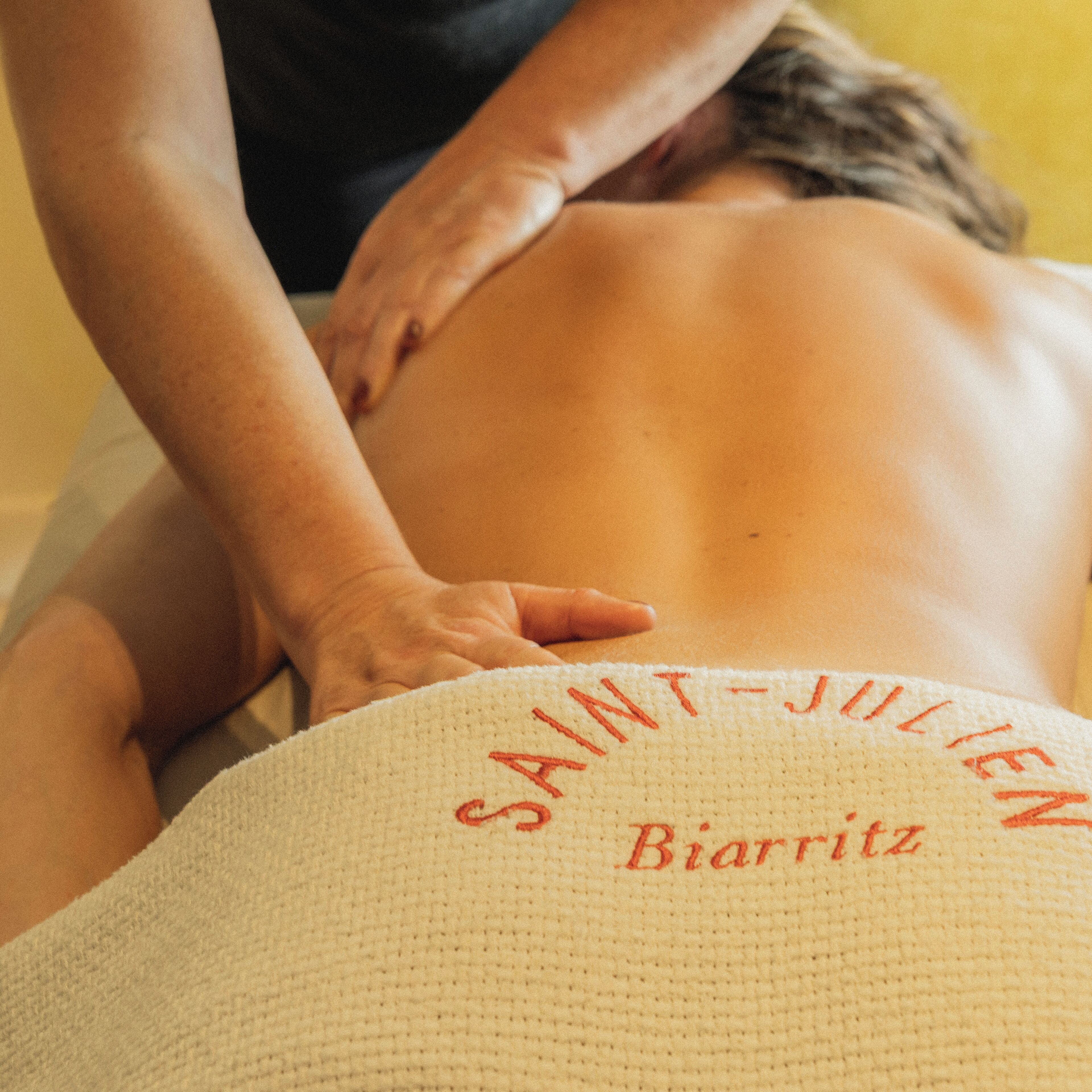 Uma pessoa recebendo uma massagem nas costas, com uma toalha com a inscrição "SAINT-JULIEN Biarritz" no chão.