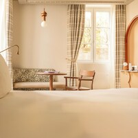 Um quarto aconchegante com uma cama, uma cadeira de madeira, uma mesinha com um livro e uma janela com cortinas.