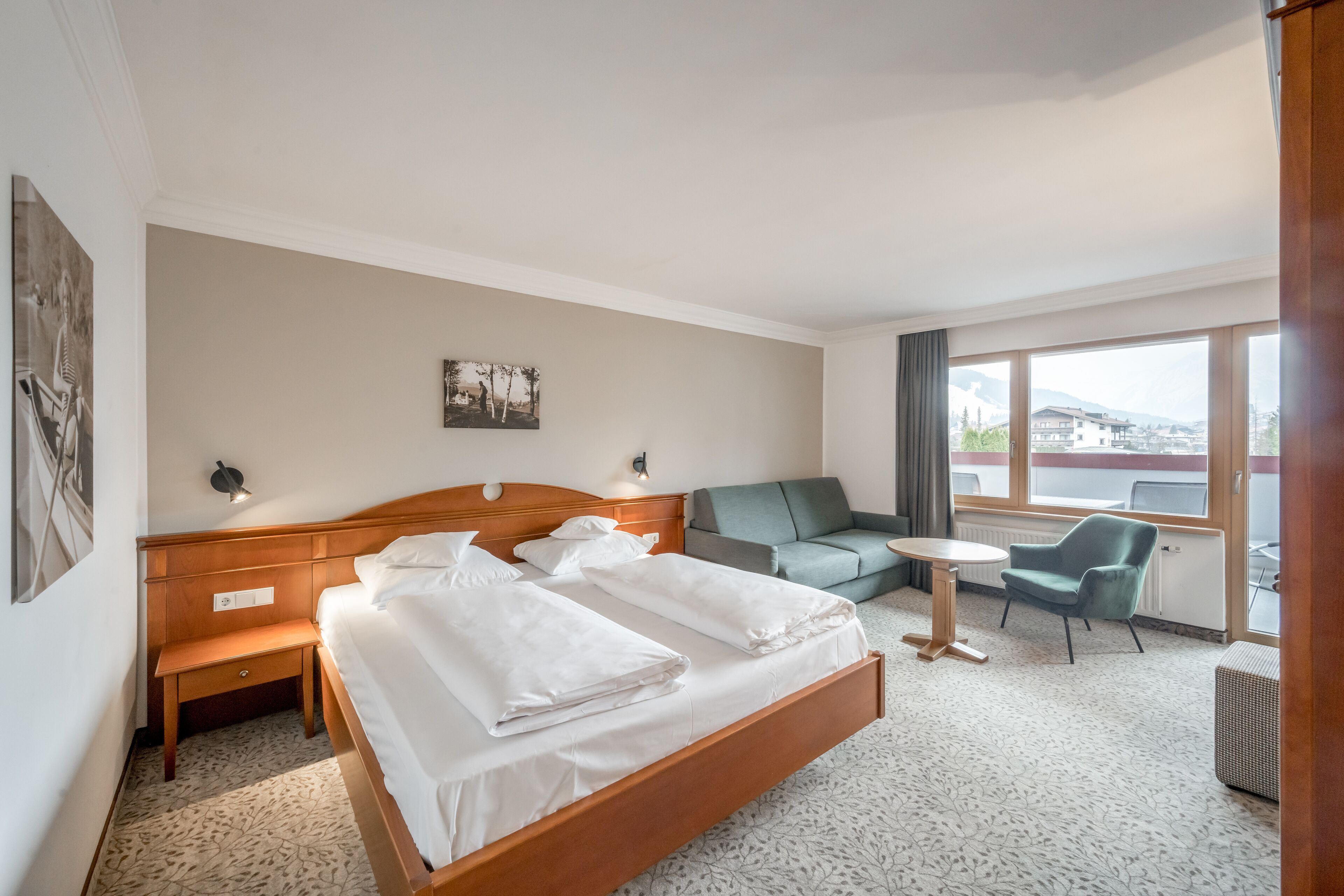 Quarto de hotel com uma cama grande, um sofá, uma mesinha e vista da cidade através de amplas janelas.