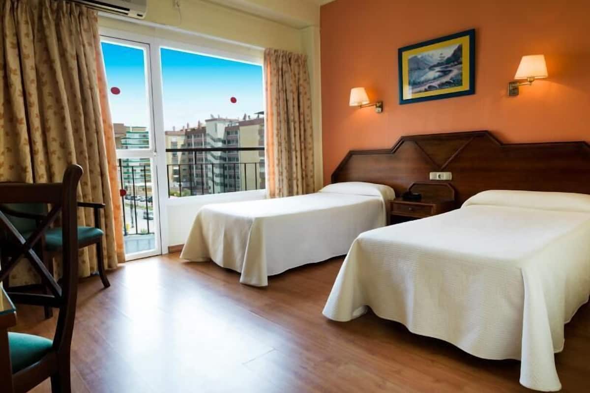 Quarto de hotel com duas camas, uma cadeira, uma mesa, uma mesinha de cabeceira e um quadro na parede.