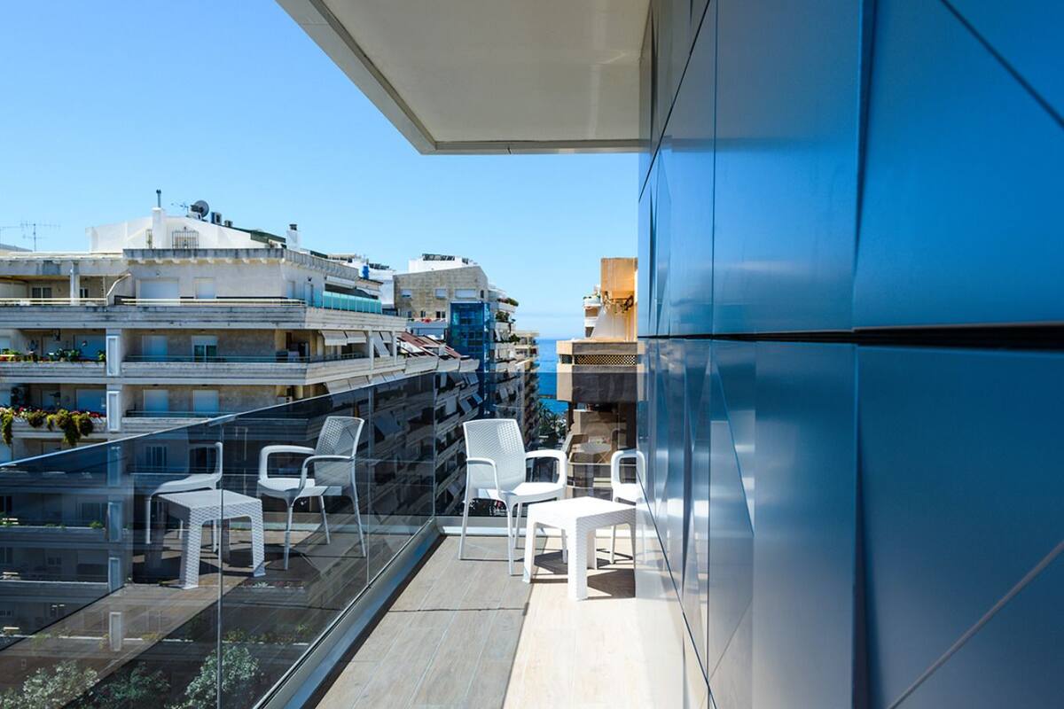 Varanda com cadeiras brancas e guarda-corpo de vidro, com vista para um horizonte urbano repleto de edifícios e céu claro.