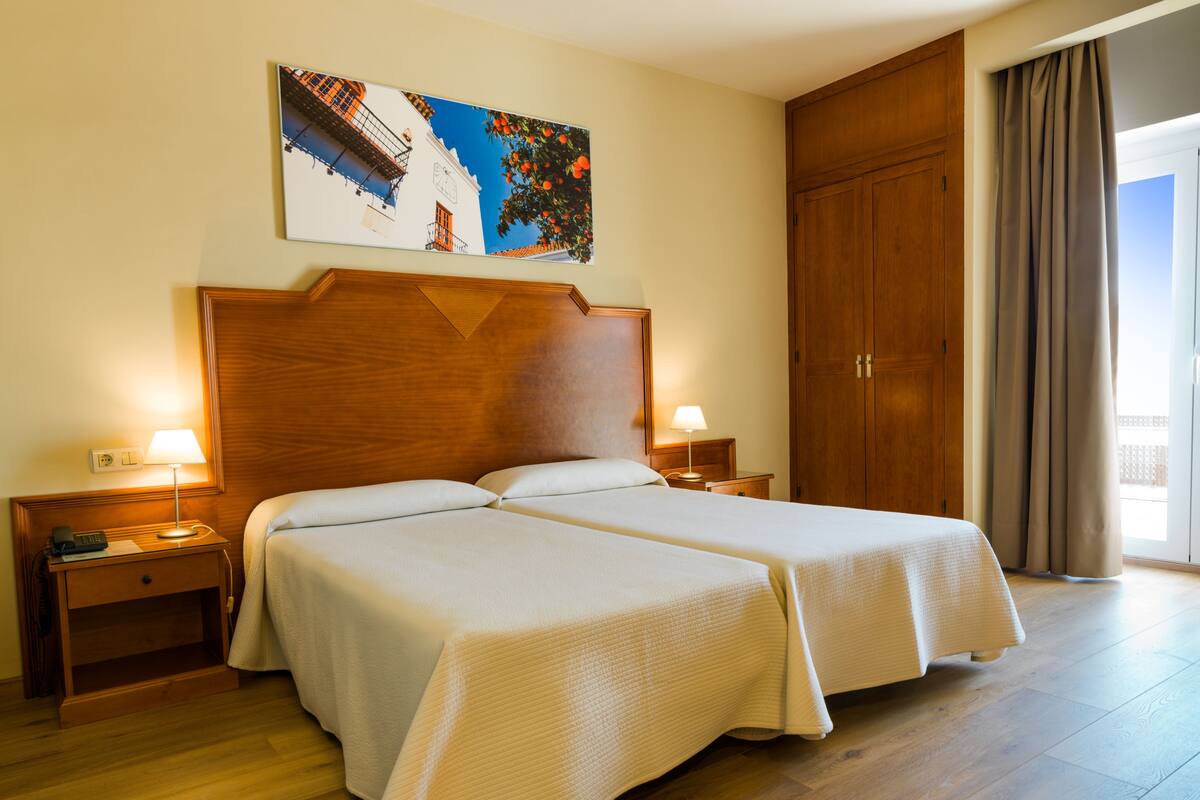 Quarto de hotel com duas camas, cabeceira de madeira, mesas de cabeceira com luminárias, um quadro na parede e um guarda-roupa espaçoso.