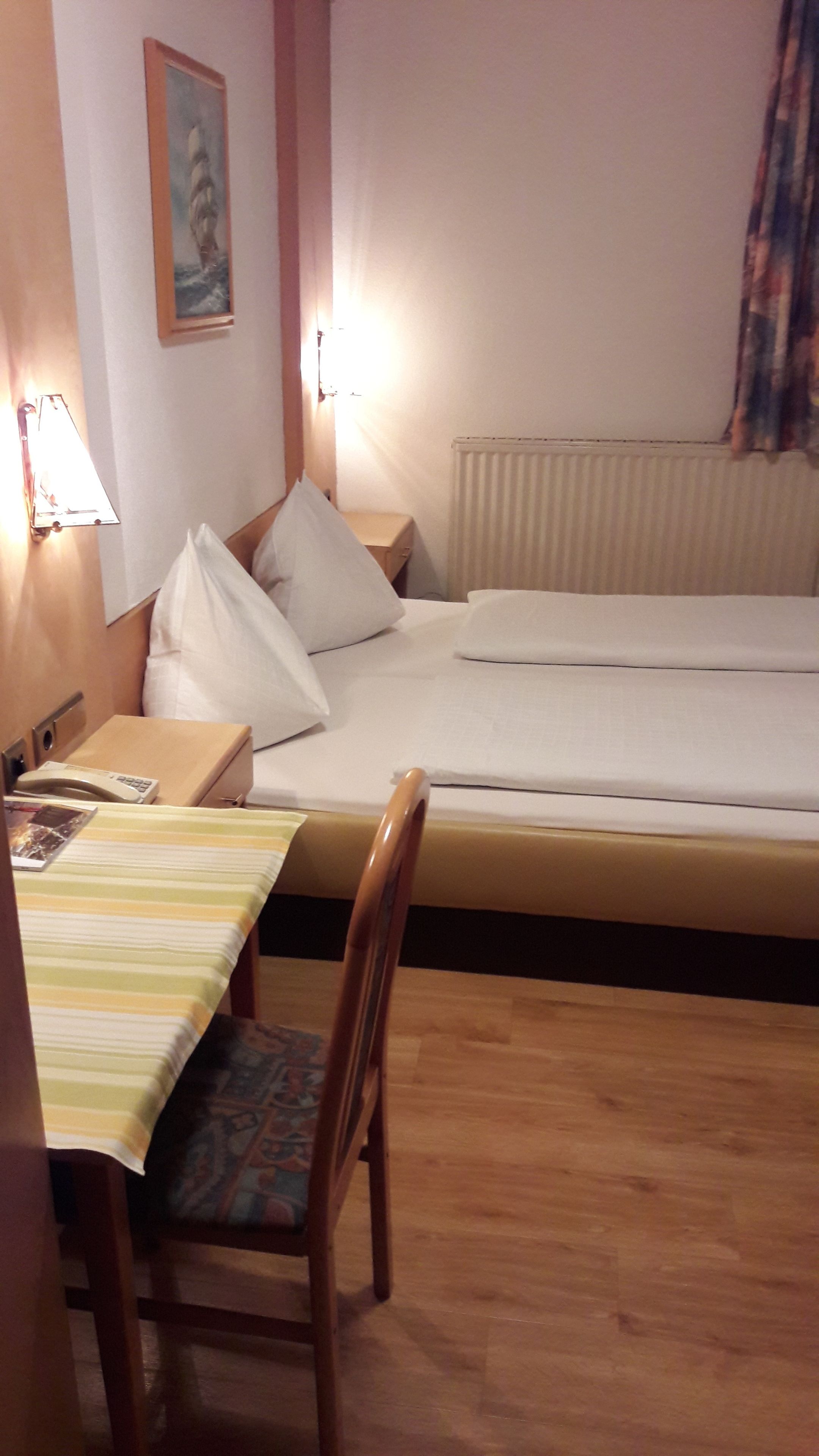 Quarto de hotel com cama, escrivaninha, cadeira, abajur e um quadro na parede.
