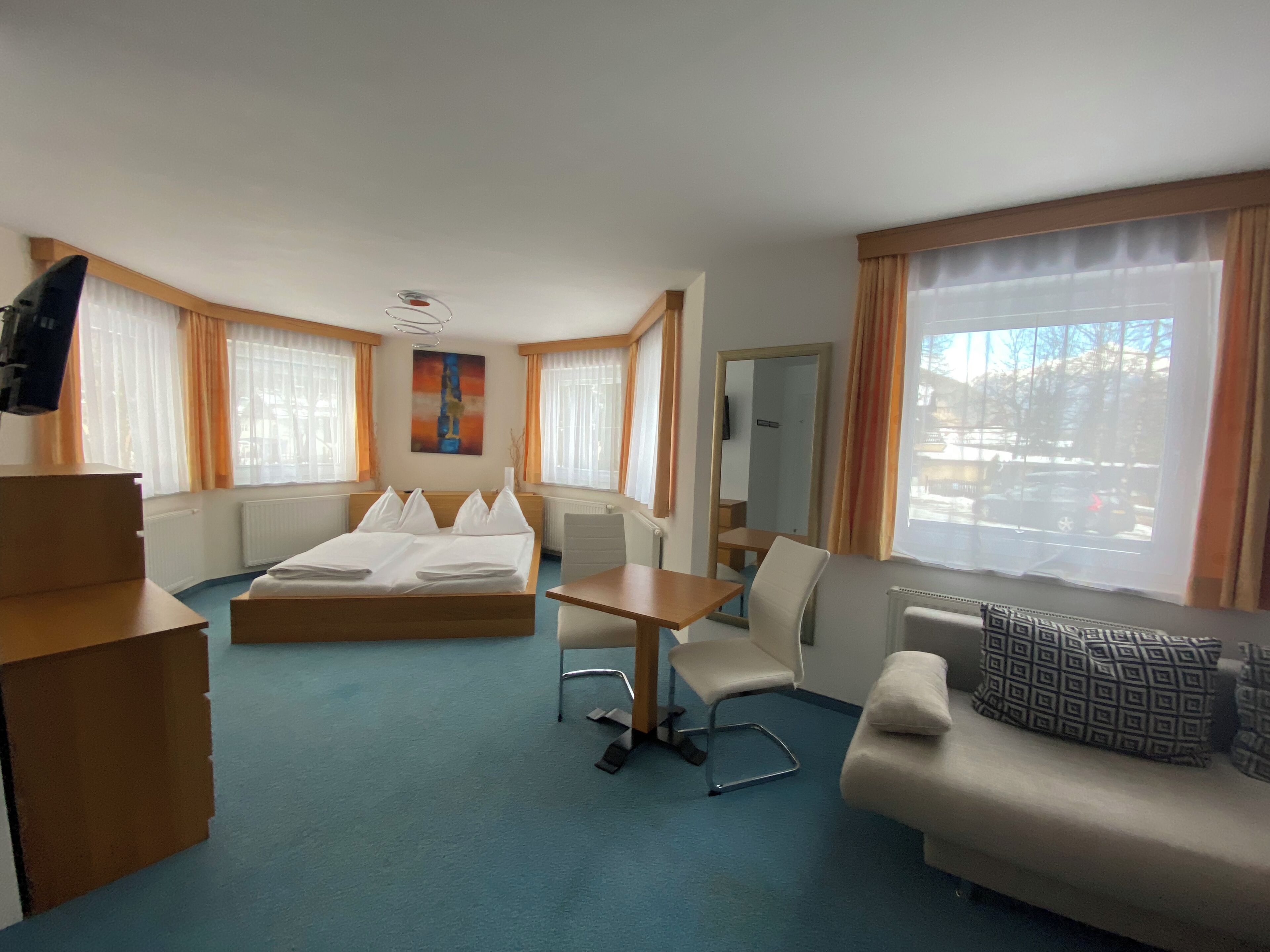 Quarto de hotel com cama, sofá, mesa pequena, cadeira, TV e janela com cortinas.