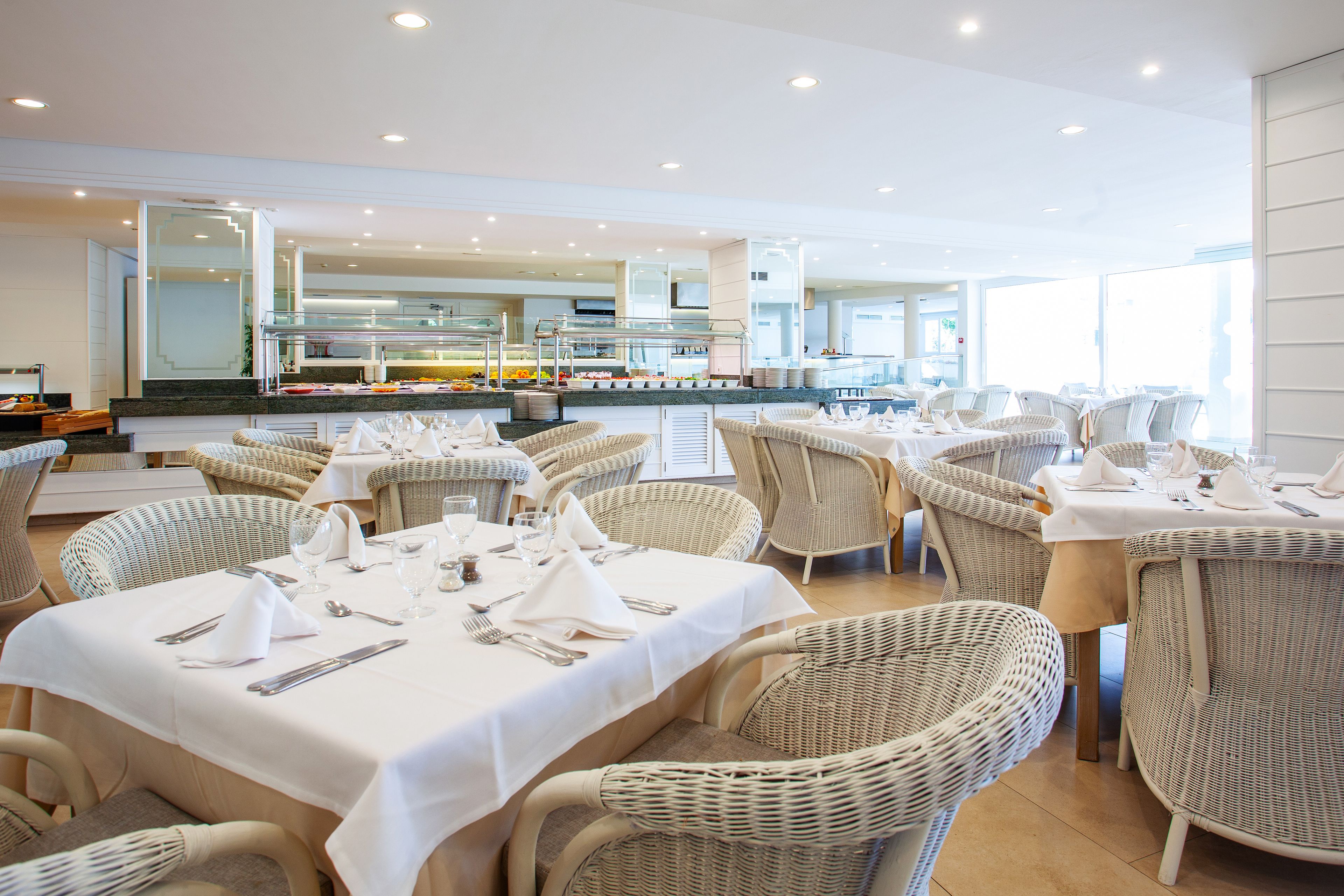 Restaurante com cadeiras e mesas de vime dispostas para refeições, área de buffet ao fundo e grandes janelas que oferecem luz natural.