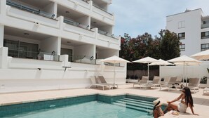 Uma área de piscina com espreguiçadeiras e guarda-sóis, piscina azul cristalina e um edifício branco com varandas.