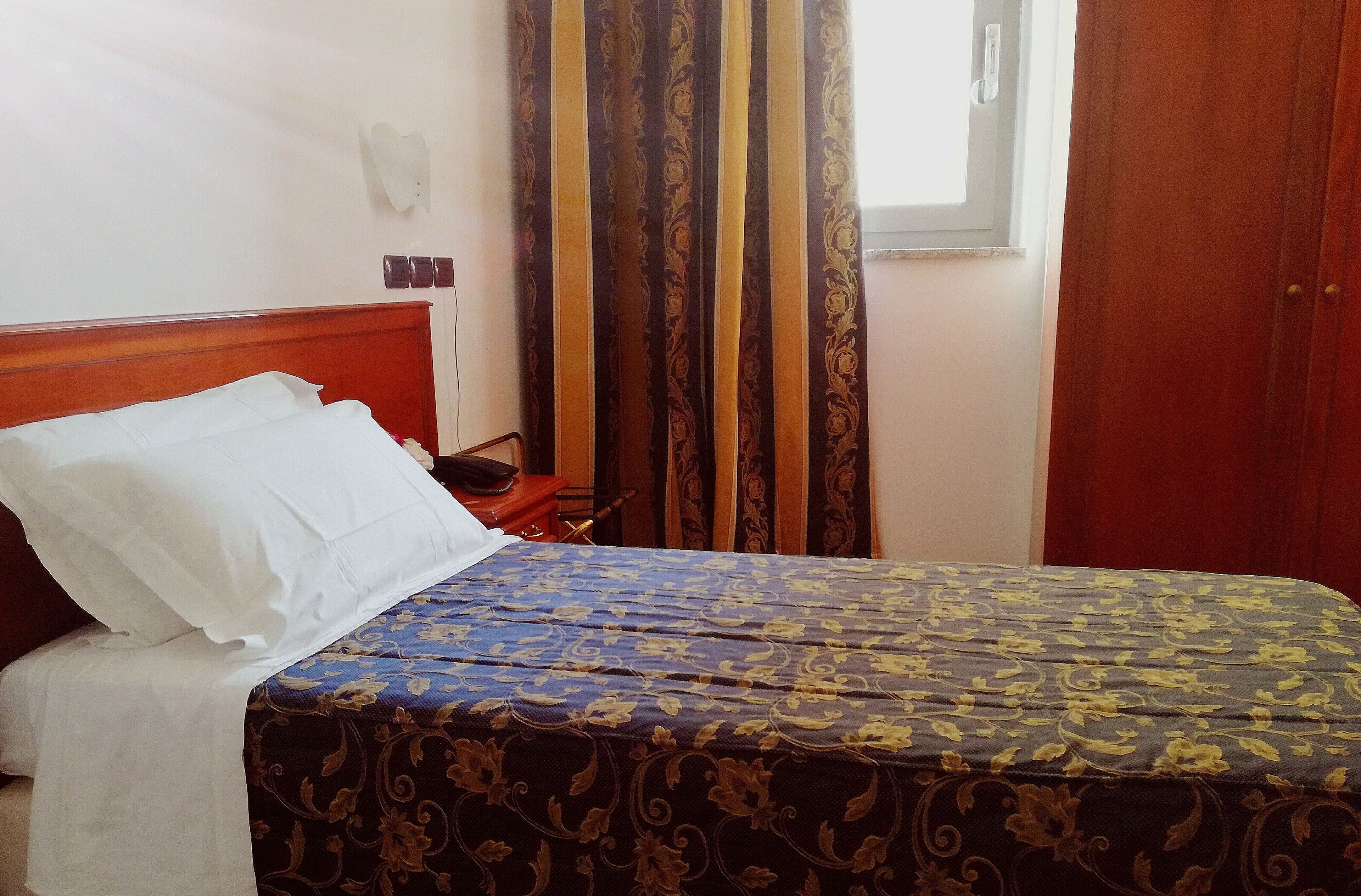 Uma cama bem arrumada com colcha florida, cabeceira de madeira e uma janela com cortinas.