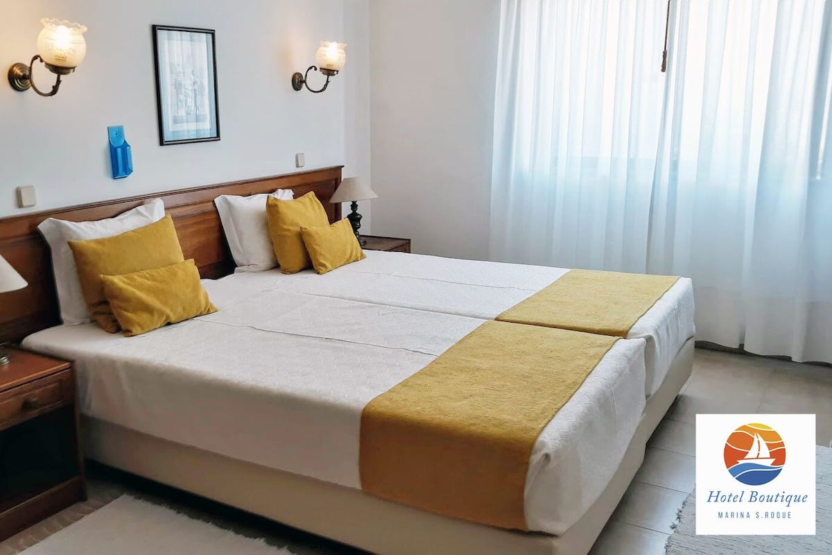 Quarto de hotel com uma cama grande, duas mesas de cabeceira, um criado-mudo, uma luminária e um quadro na parede.