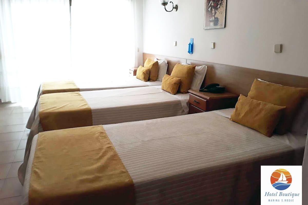 Quarto de hotel com duas camas de solteiro, cada uma com colcha bege e almofadas a combinar. Há uma mesa de cabeceira com abajur e telefone, e uma prateleira na parede com um objeto azul. Uma pintura é visível na parede, e o quarto tem uma janela com cortinas finas.