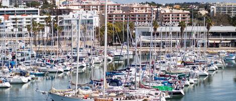 Uma marina com vários barcos atracados, um edifício com a inscrição "MARINA DE LAGOS" ao fundo e uma paisagem urbana com edifícios residenciais.