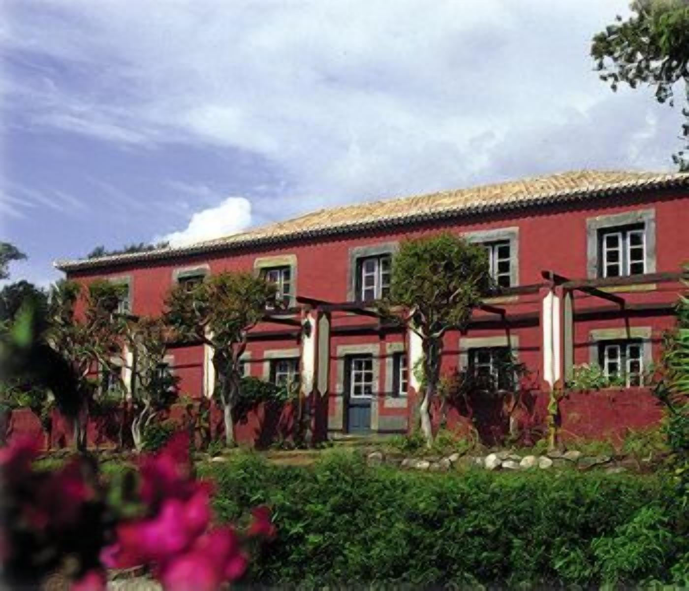 Um edifício vermelho com telhado de telhas, rodeado por vegetação e árvores.