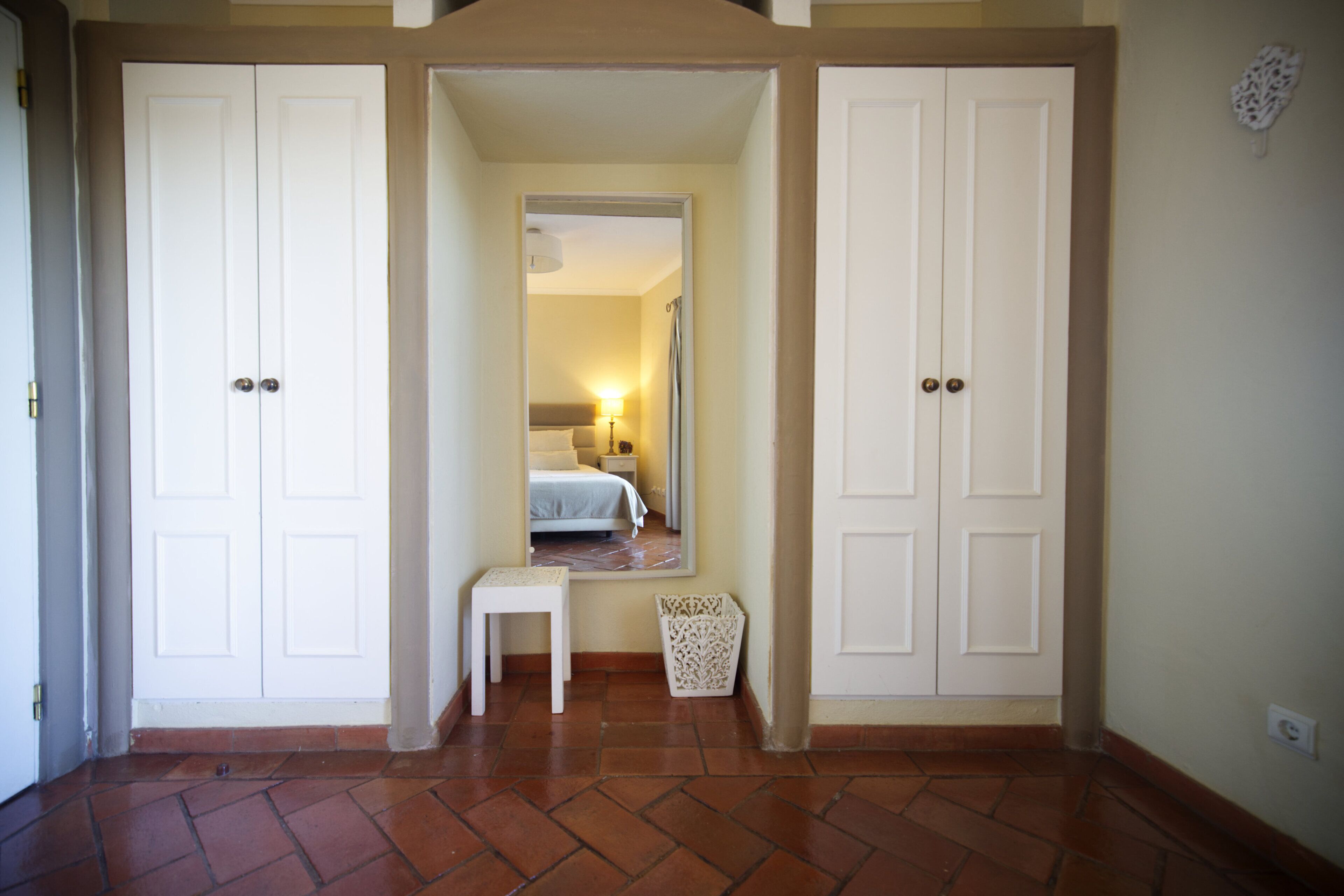 Um corredor com duas portas brancas, uma pequena mesa branca e um espelho refletindo um quarto com uma cama e mesa de cabeceira.