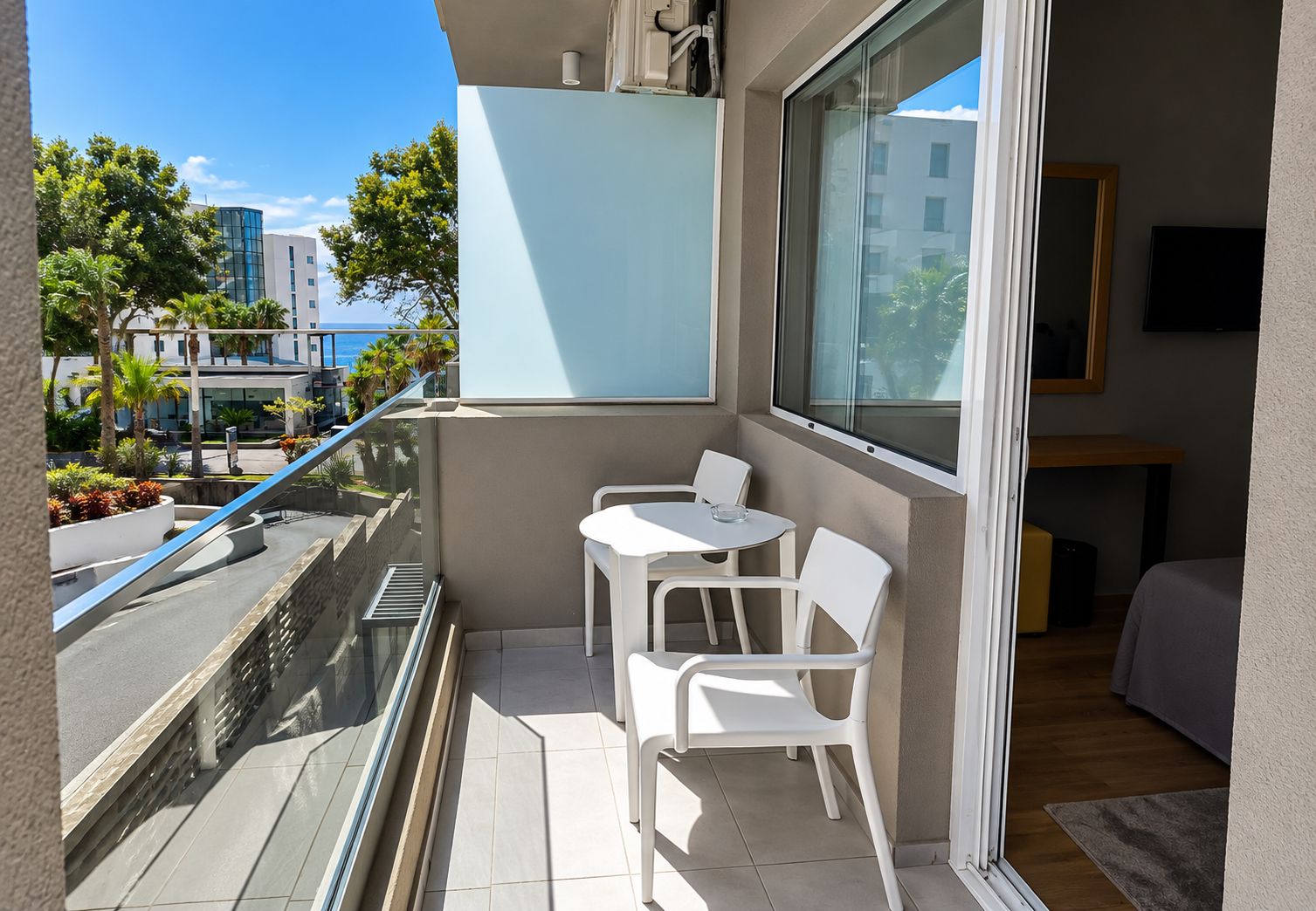 Uma varanda com guarda-corpo de vidro, uma mesa pequena com duas cadeiras e vista para o mar e edifícios.