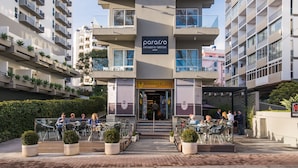 Um edifício moderno com um letreiro indicando 'paratro apartments' e 'supermercado'. Há áreas de estar ao ar livre com mesas e cadeiras, e uma escadaria que leva à entrada.