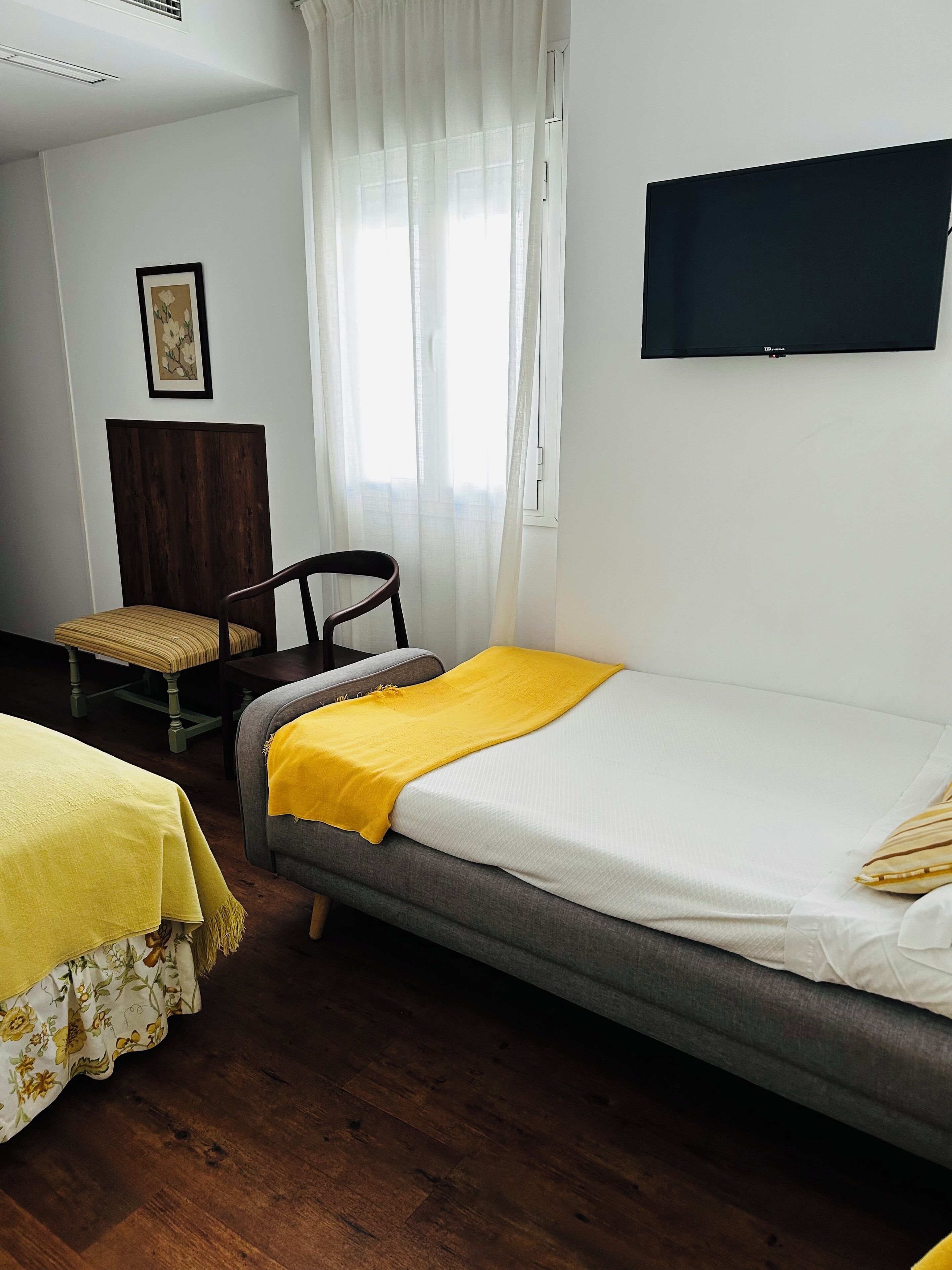 Quarto de hotel com cama, cadeira, escrivaninha, televisão e janela com cortinas.