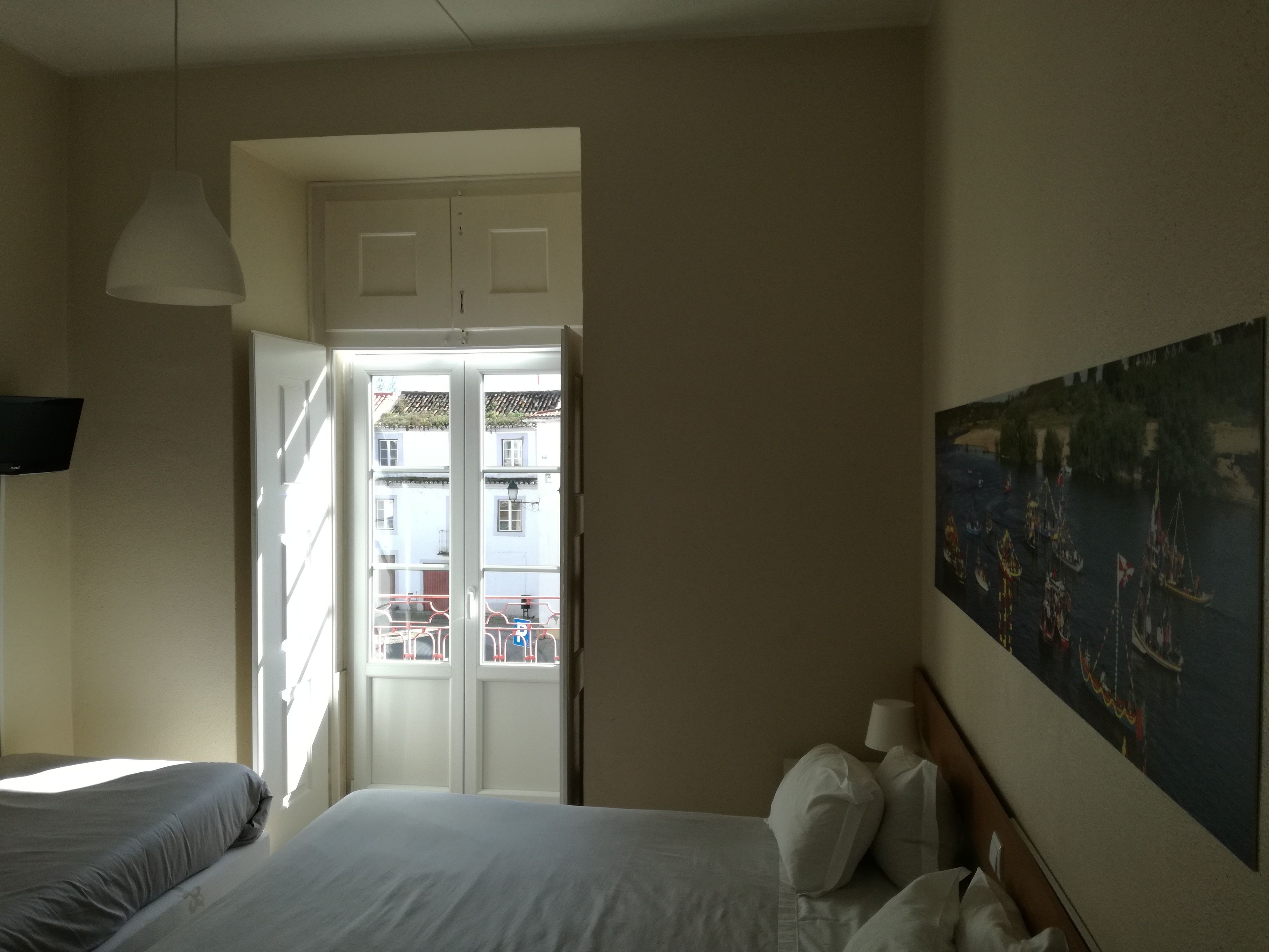 Quarto de hotel com uma cama, um quadro na parede e vista para edifícios através de uma porta.