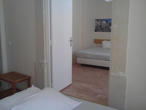 Quarto pequeno e limpo, com uma cama, um banquinho de madeira e uma porta que dá para outro espaço.